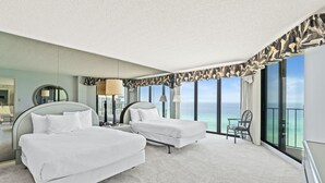 Quarto de hotel com duas camas, uma janela grande com vista para o oceano e uma varanda com uma cadeira.