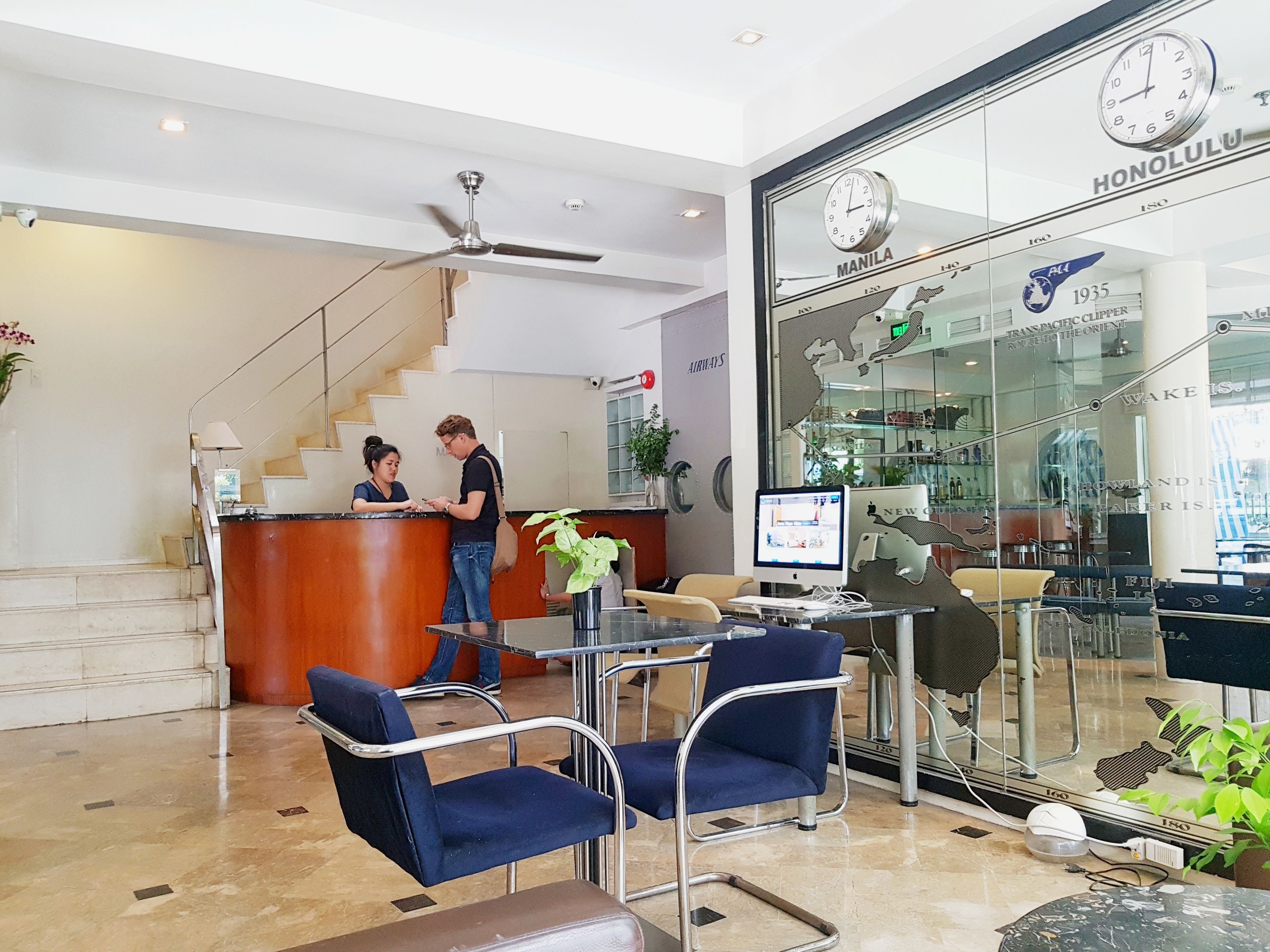 Uma área de recepção de hotel com balcão, tela digital, espaço para sentar e uma escadaria.