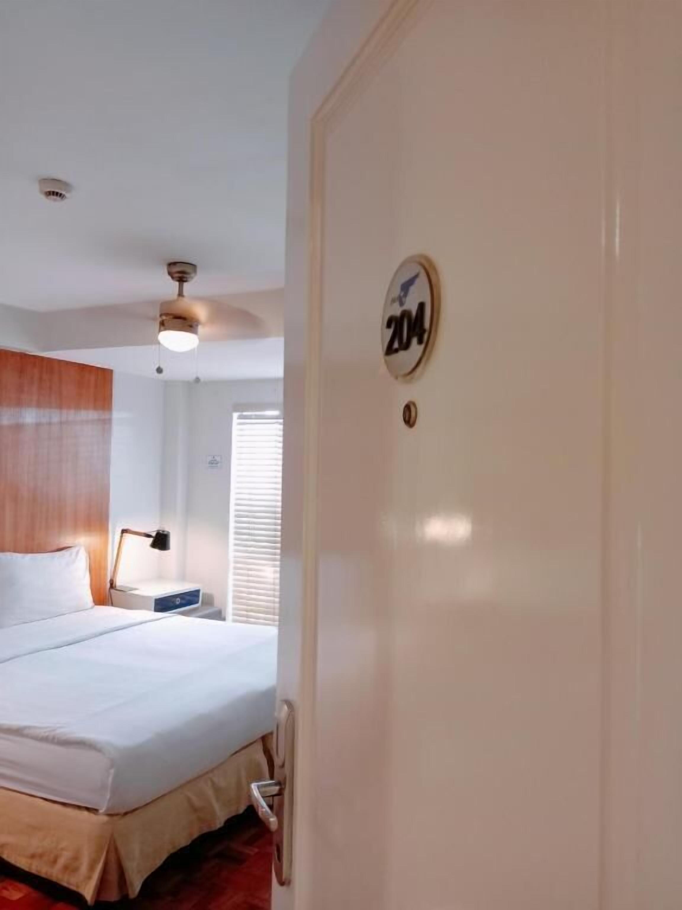 Quarto de hotel com cama, mesa de cabeceira com luminária, ventoinha no teto e porta com número do quarto.