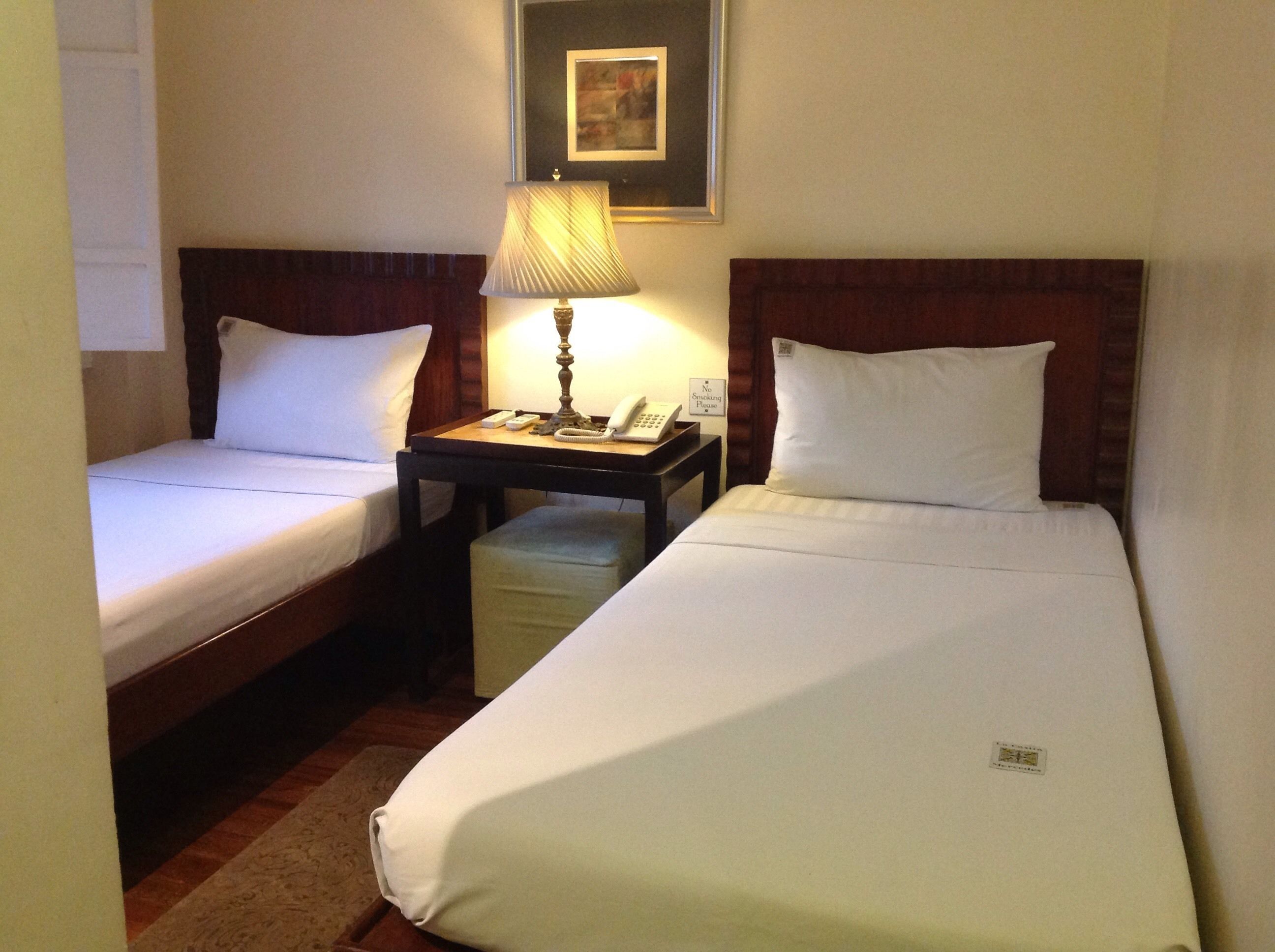 Quarto de hotel com duas camas de solteiro, uma mesa de cabeceira com abajur, telefone e placa indicando 'Proibido Fumar'.