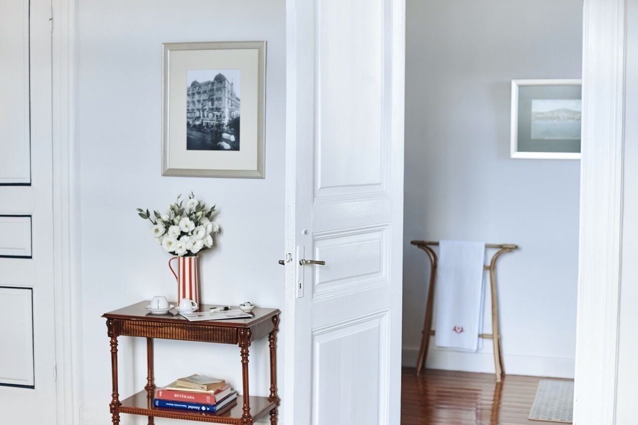 Quarto com uma mesinha de madeira com livros e um vaso de flores, uma fotografia a preto e branco emoldurada na parede, e uma porta branca que dá para outro quarto.