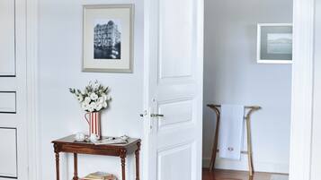 Quarto com uma mesinha de madeira com livros e um vaso de flores, uma fotografia a preto e branco emoldurada na parede, e uma porta branca que dá para outro quarto.