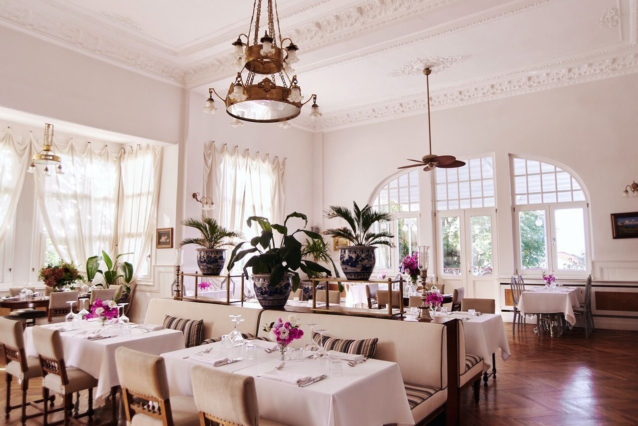 Uma área de refeições com mesas preparadas para o serviço, grandes janelas e plantas decorativas.