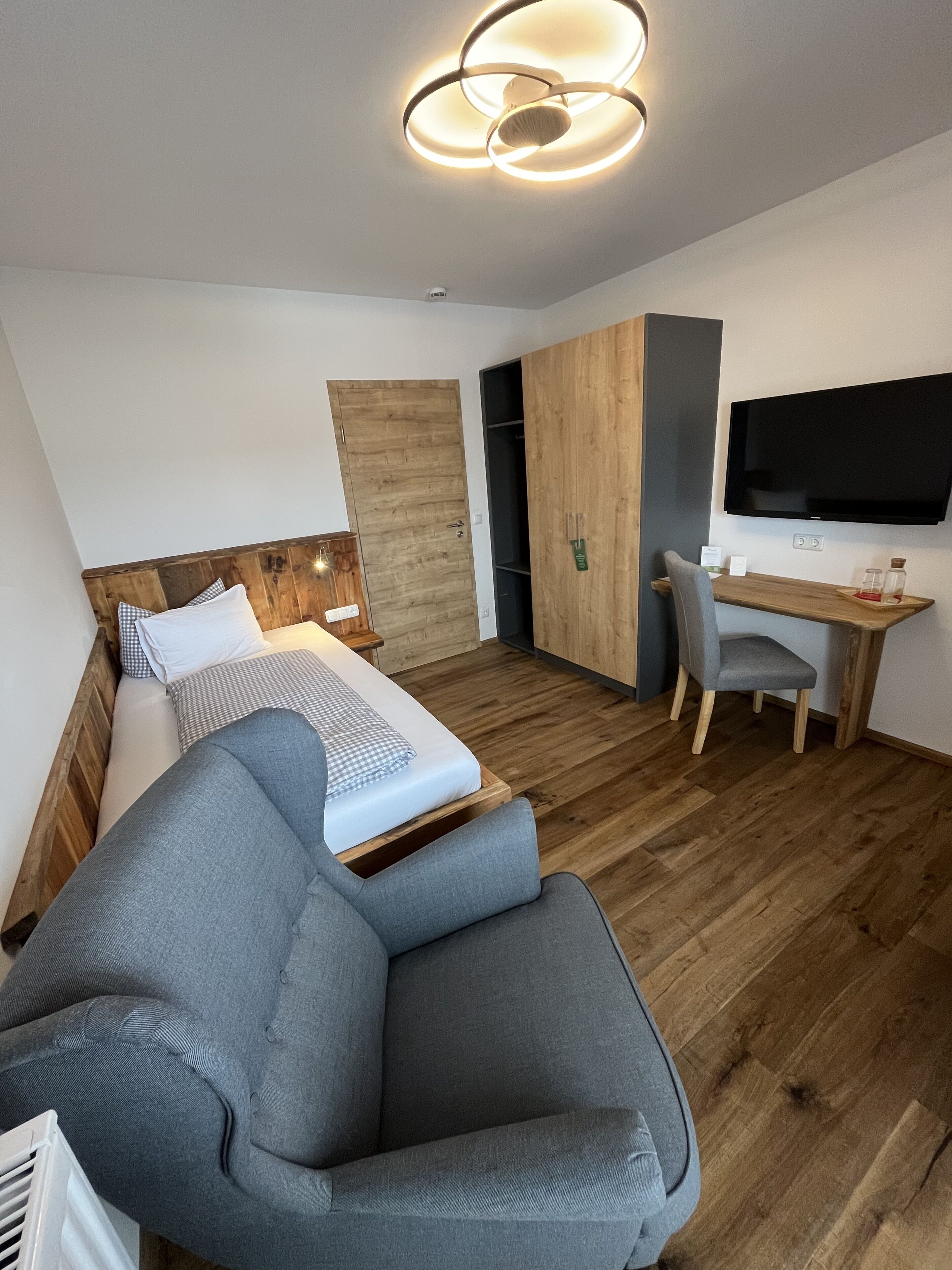 Quarto de hotel moderno com uma cama, um sofá cinzento, uma secretária de madeira, uma televisão e um guarda-roupa de madeira.