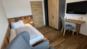 Quarto de hotel moderno com uma cama, um sofá cinzento, uma secretária de madeira, uma televisão e um guarda-roupa de madeira.