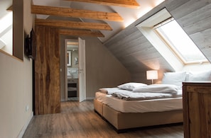 Quarto com teto inclinado, vigas de madeira, claraboia, cama com roupa de cama branca e uma mesa de cabeceira.