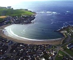 Vista aérea de uma vila costeira com uma praia curva, áreas residenciais e um pequeno porto.