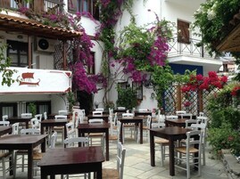 Café ao ar livre com mesas e cadeiras, rodeado por vegetação exuberante e plantas floridas.