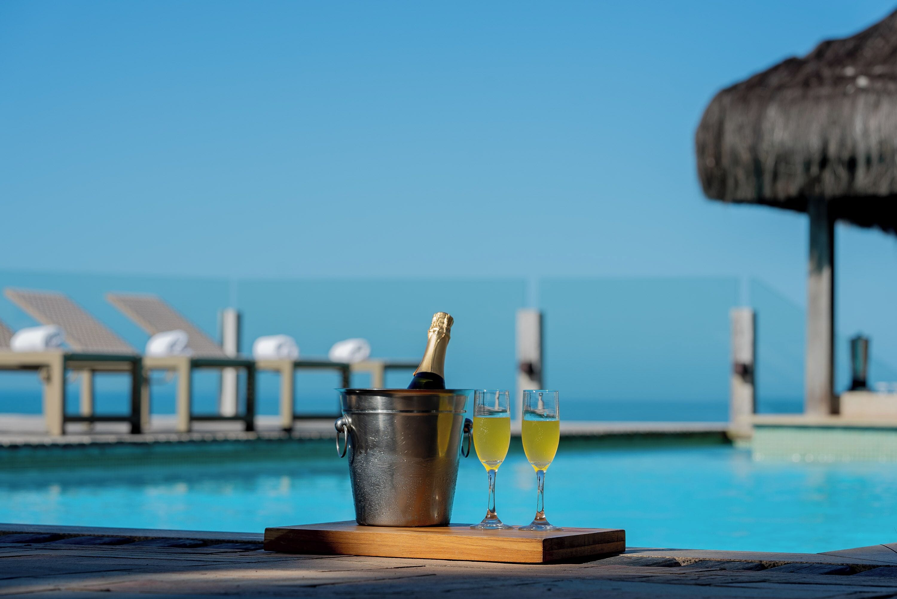 Cenário à beira da piscina, com um balde de metal contendo uma garrafa de champanhe e duas taças, apoiado em uma tábua de madeira.