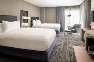 Quarto de hotel com duas camas, uma escrivaninha, uma cadeira, uma televisão e uma janela grande com cortinas.
