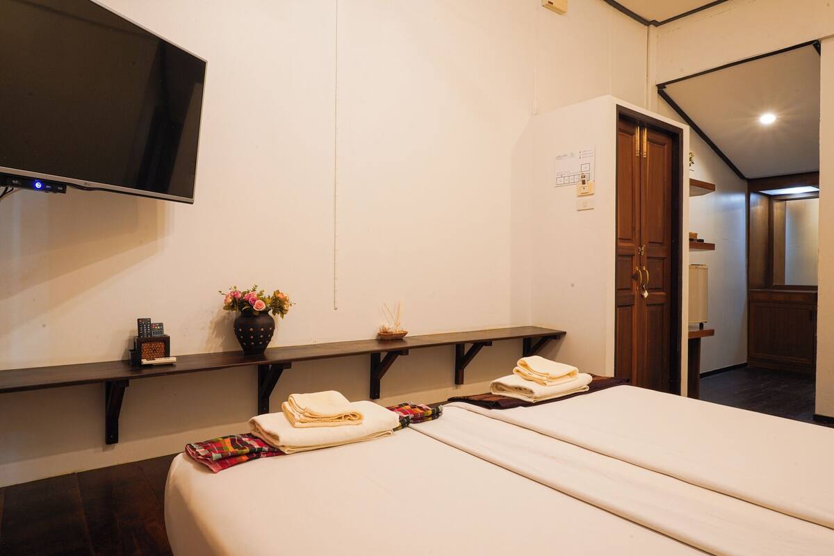 Quarto de hotel com uma cama, televisão fixada na parede, uma mesinha com um vaso de flores e uma porta de madeira.