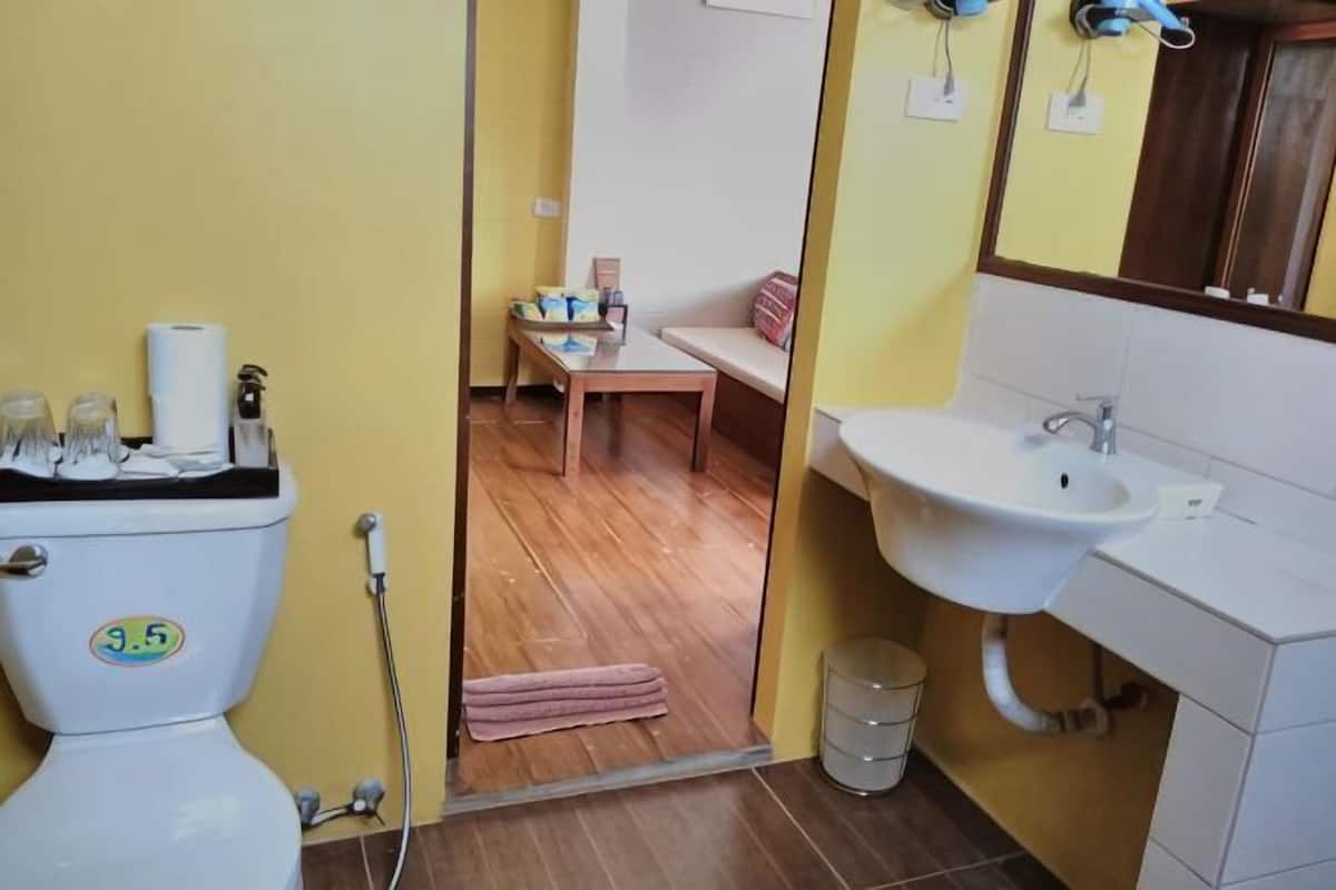 Casa de banho com sanita, lavatório e espelho. Tem chão de madeira e uma pequena mesa com objetos sobre ela.