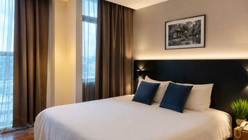 Quarto de hotel com uma cama grande, cabeceira com iluminação integrada, quadro na parede e vista da cidade pela janela.