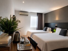 Quarto de hotel com duas camas, uma mesinha de cabeceira com chaleira, um suporte com garrafas de água e uma planta em vaso.