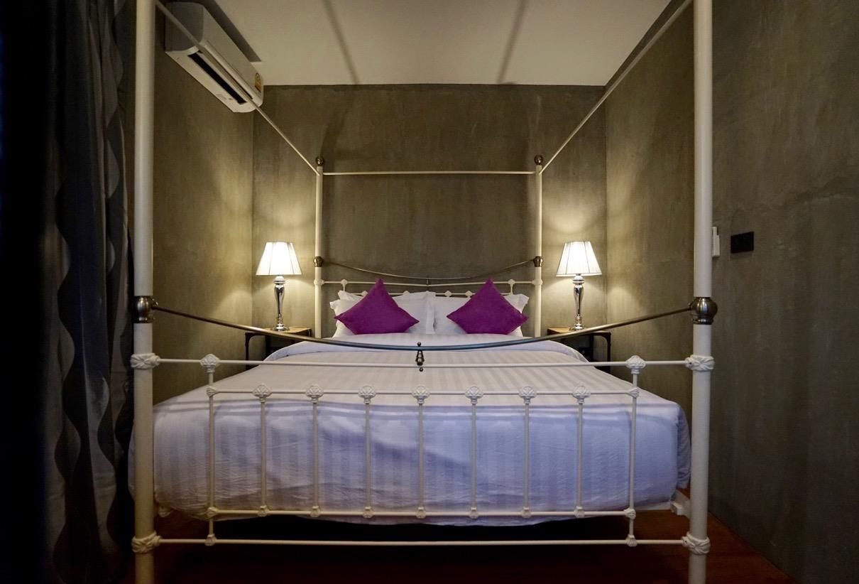 Uma cama de dossel com estrutura metálica branca e colcha branca, dois travesseiros roxos e duas luminárias de cabeceira.