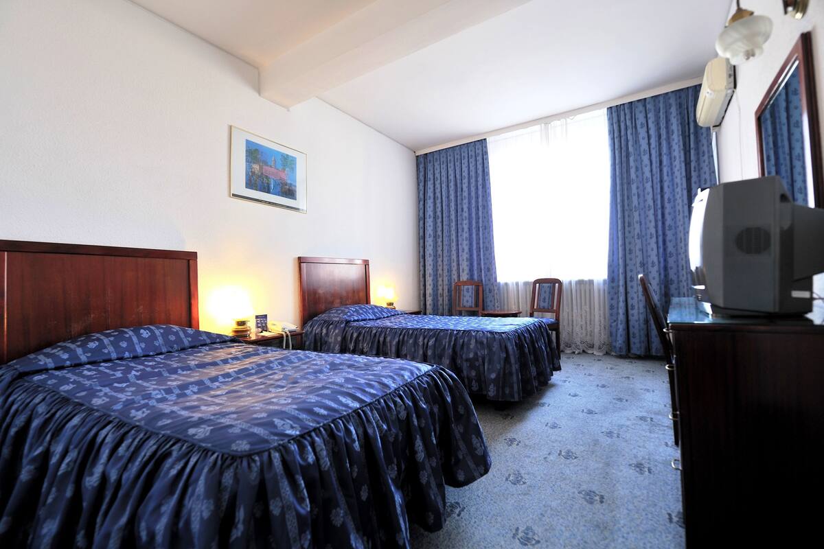 Quarto de hotel com duas camas, uma escrivaninha, uma cadeira, uma televisão e uma janela com cortinas.