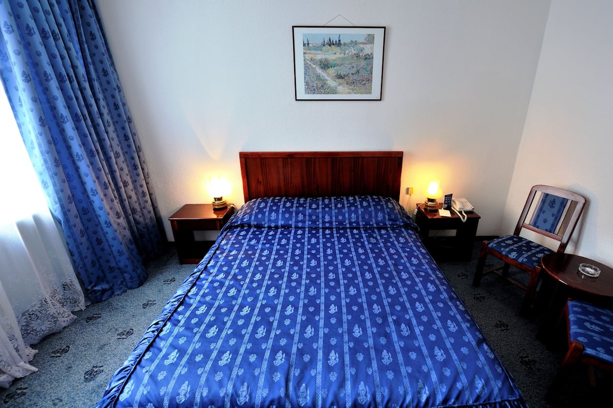Quarto de hotel com uma cama grande, duas mesas de cabeceira com luminárias, um quadro na parede e uma poltrona com almofada.