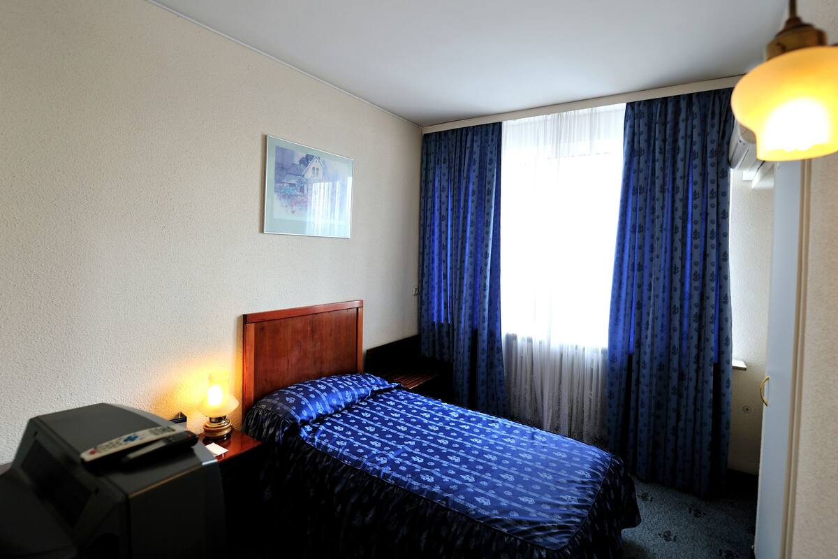 Quarto de hotel com uma cama, uma escrivaninha com luminária, um quadro na parede e uma janela com cortinas.