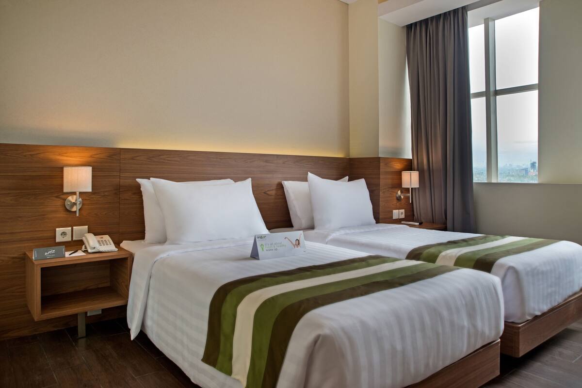 Quarto de hotel com duas camas, cabeceira de madeira, luminárias de cabeceira, telefone e janela com cortinas.