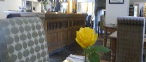 Uma sala de jantar com uma mesa de madeira, uma cadeira com almofada de bolinhas e um vaso com uma rosa amarela.