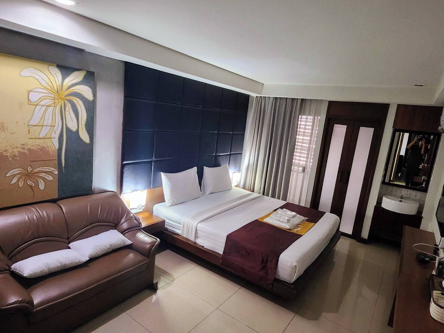 Quarto de hotel com uma cama grande, um sofá de couro marrom, uma mesinha de cabeceira, uma parede com uma pintura de palmeira, uma janela com cortinas e um espelho com uma pia.