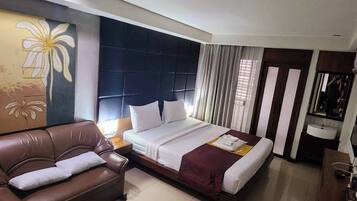 Quarto de hotel com uma cama grande, um sofá de couro marrom, uma mesinha de cabeceira, uma parede com uma pintura de palmeira, uma janela com cortinas e um espelho com uma pia.