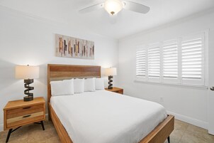 Quarto com cama de madeira, roupa de cama branca, duas mesas de cabeceira com luminárias, ventoinha no teto, janela com persianas e um quadro abstrato pendurado na parede.
