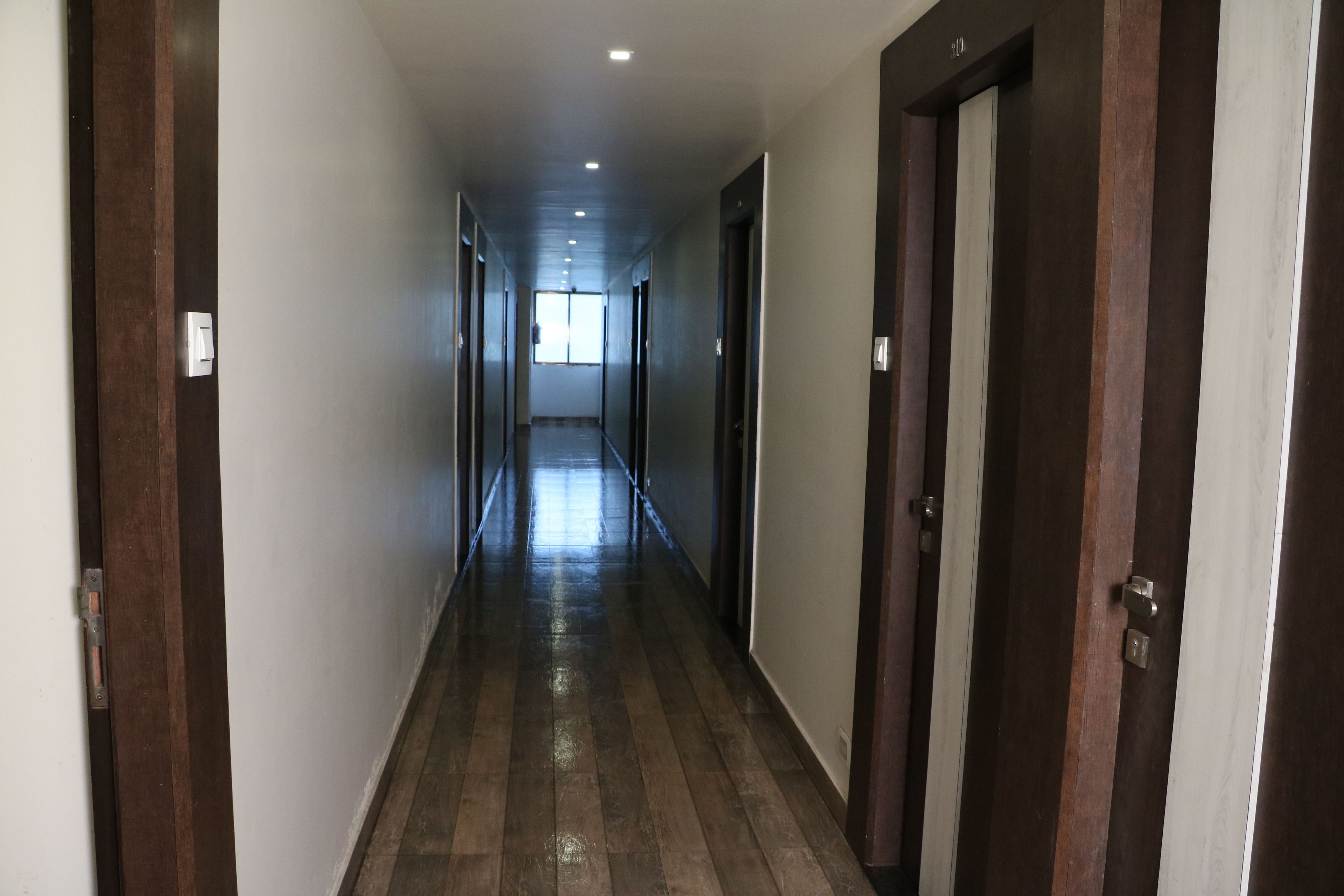 Um corredor comprido e estreito com portas de madeira em ambos os lados, dando para um quarto iluminado no final.