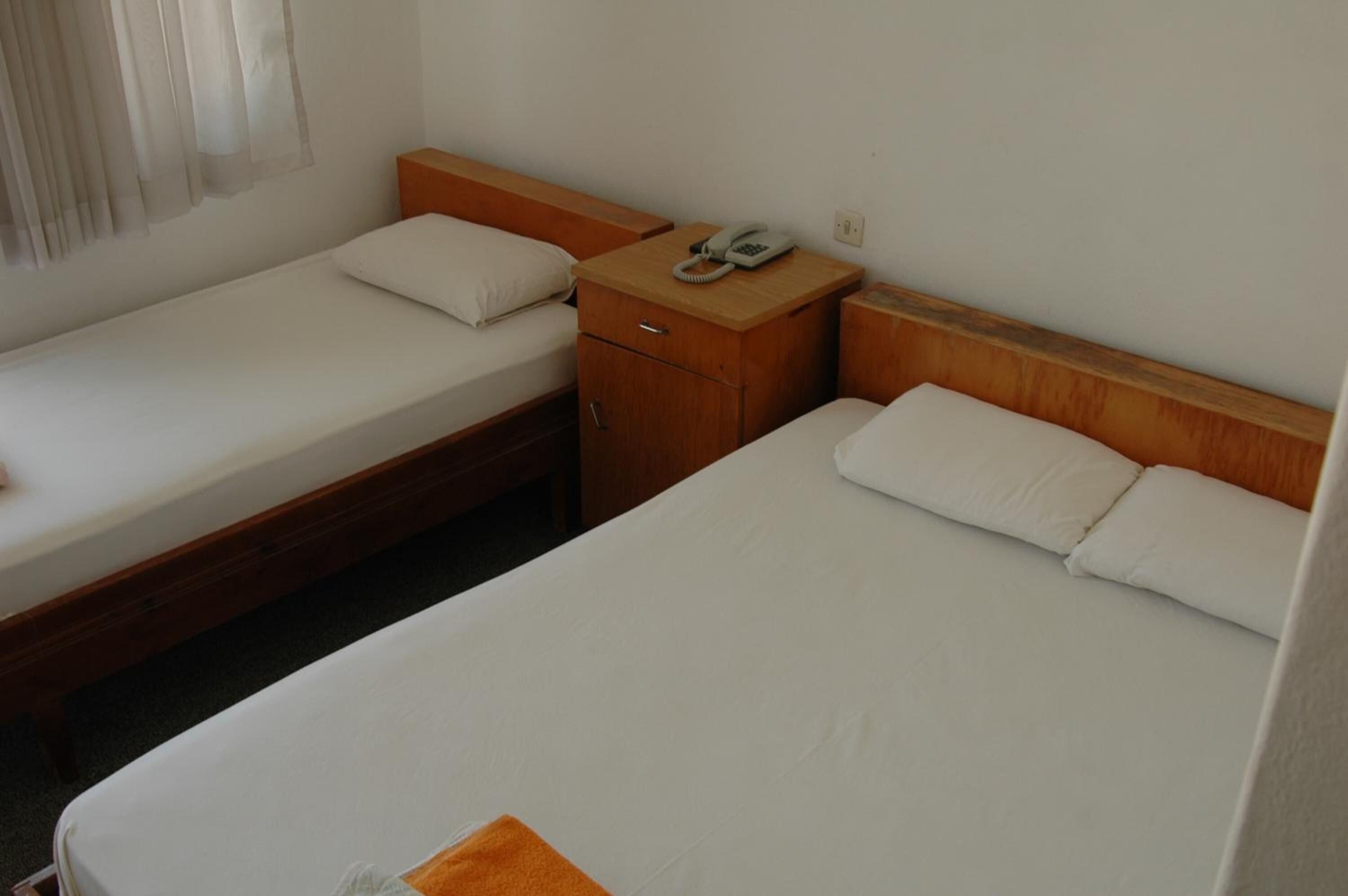 Quarto de hotel com duas camas de solteiro, mesa de cabeceira de madeira com telefone e uma pequena gaveta.