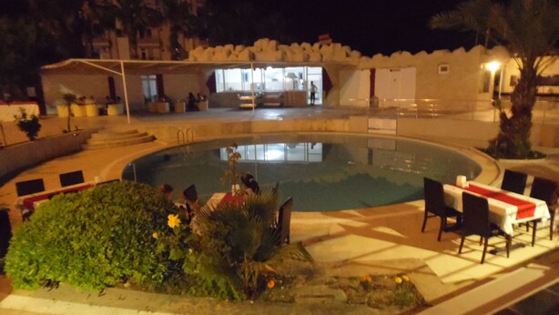 Área de piscina à noite, com espaço para refeições e um edifício ao fundo.