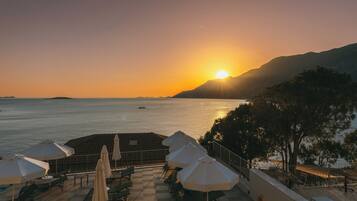 Terraço com área de estar ao ar livre e guarda-sóis, com vista para um corpo d'água ao pôr do sol.