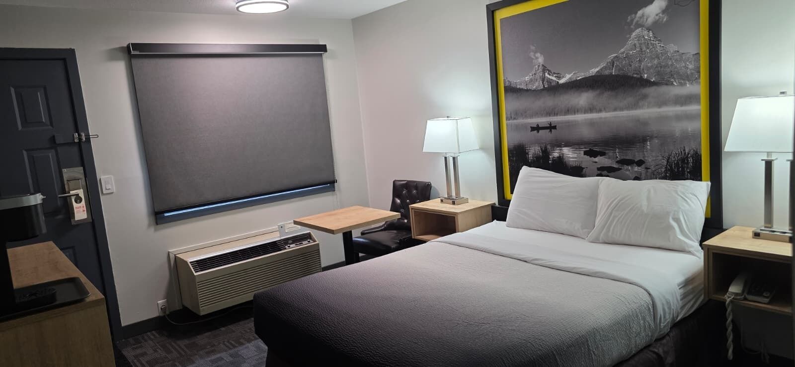 Quarto de hotel com uma cama, uma escrivaninha, uma cadeira, um aquecedor, uma janela com persiana e um candeeiro fixado na parede.