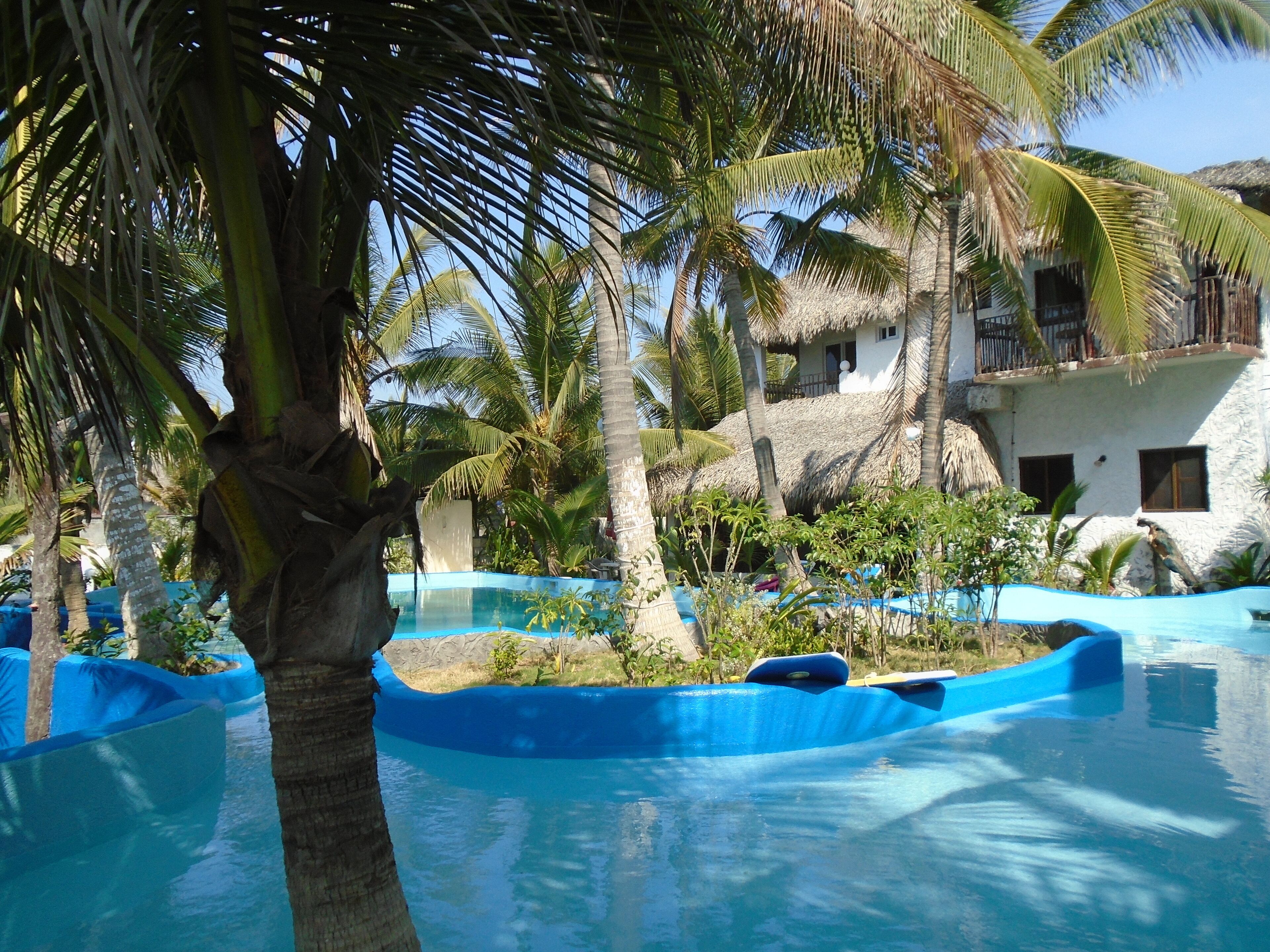 Área de piscina com palmeiras e um edifício com telhado de palha.