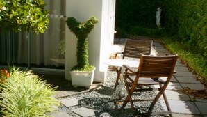 Um pátio com duas cadeiras de madeira e uma mesa, cercado por vasos de plantas e um viveiro.