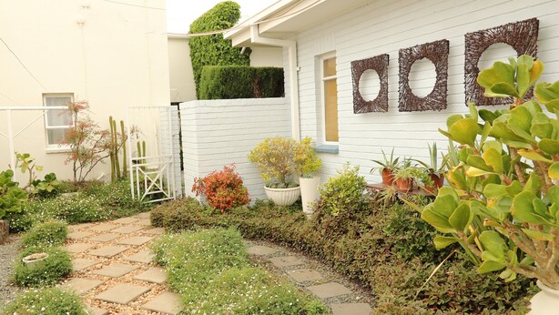 Caminho de jardim que leva a uma casa branca com decoração na parede, plantas em vasos e diversos arbustos.