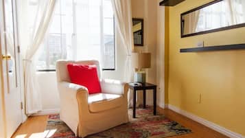 Um quarto aconchegante com uma poltrona bege, uma mesinha com abajur e um tapete colorido sobre piso de madeira.