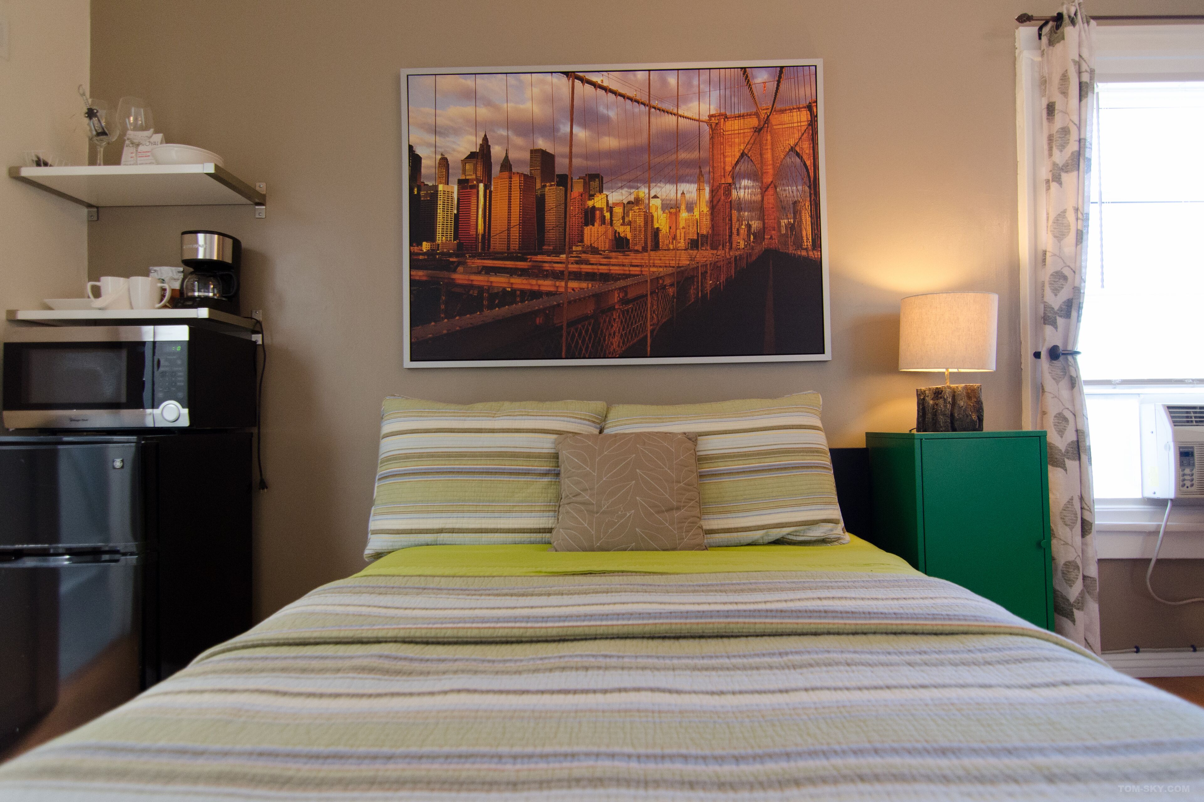 Quarto de hotel com uma cama, uma pintura de paisagem urbana, micro-ondas, frigorífico, um armário verde e uma janela com cortinas.