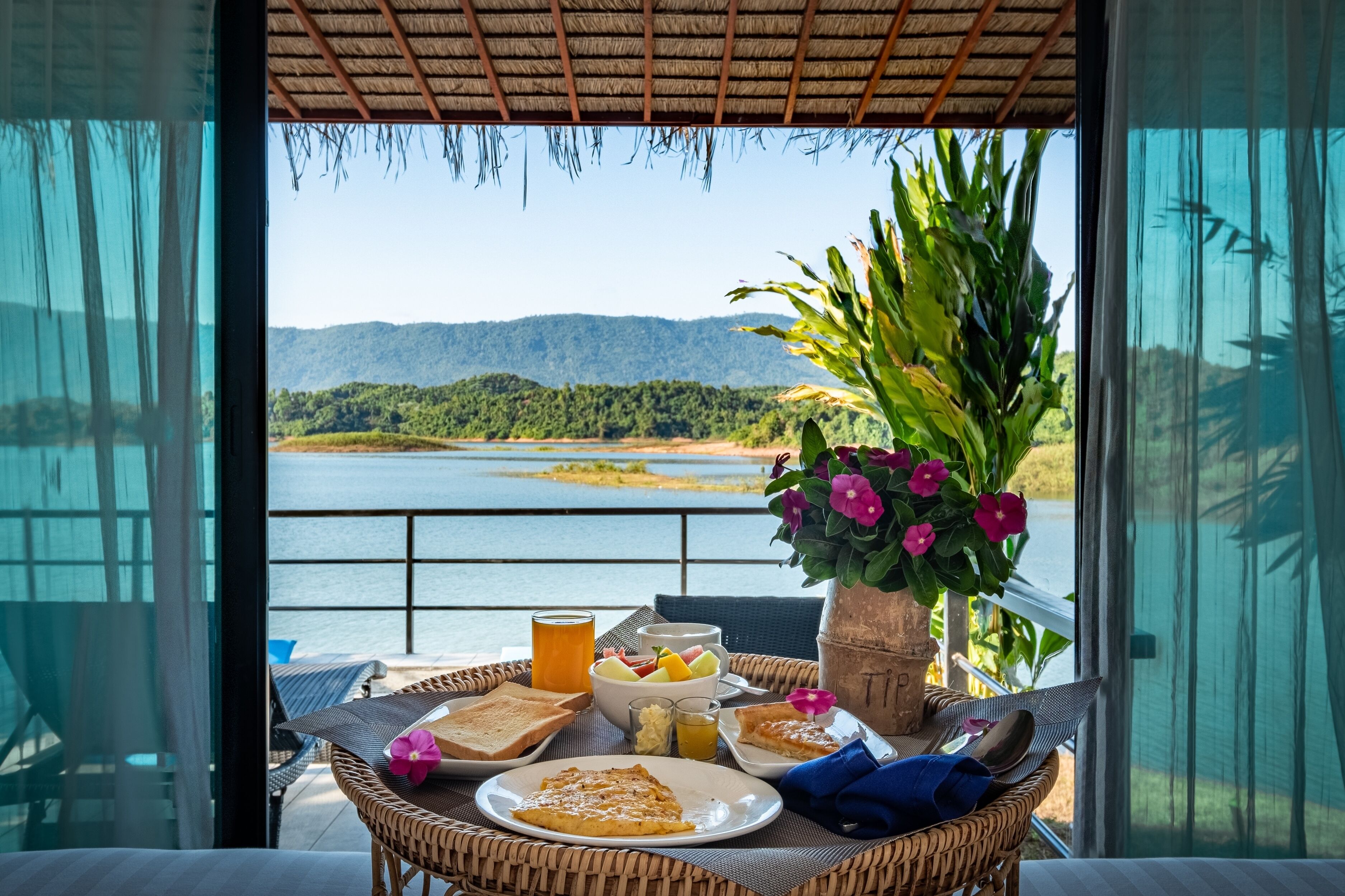 Disposição para o pequeno-almoço com vista para um lago e montanhas, incluindo uma mesa de vime, um vaso de flores e diversas opções para o café da manhã.