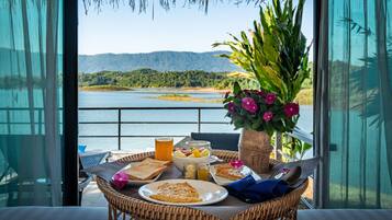 Disposição para o pequeno-almoço com vista para um lago e montanhas, incluindo uma mesa de vime, um vaso de flores e diversas opções para o café da manhã.