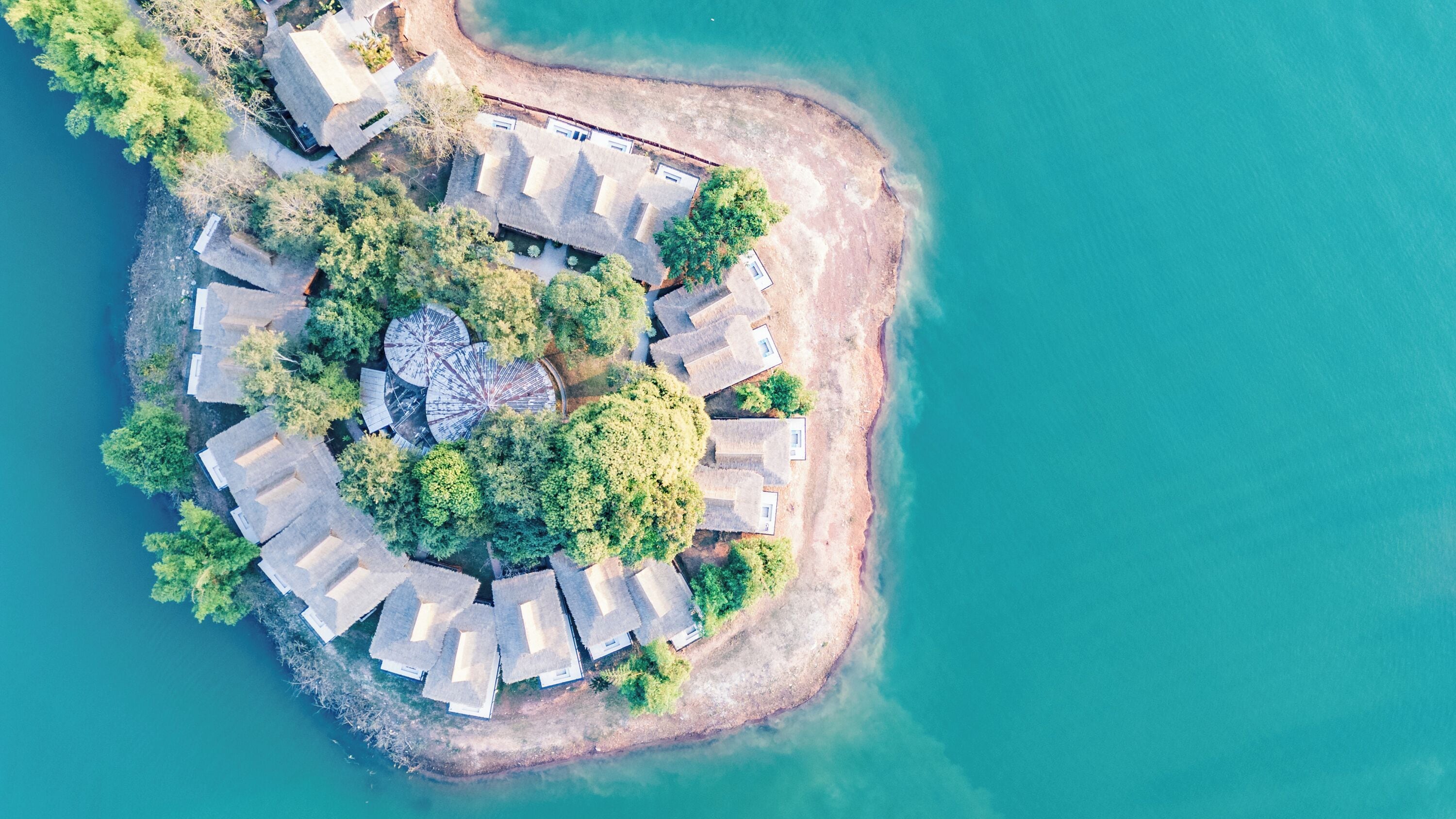 Vista aérea de um resort com chalés de telhado de palha dispostos em um padrão semicircular ao redor de uma estrutura circular central, possivelmente uma piscina ou spa, com uma praia arenosa e água turquesa cristalina.