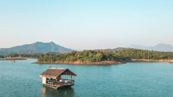 Uma casa flutuante com telhado de palha e varanda, rodeada por águas calmas e montanhas ao longe.
