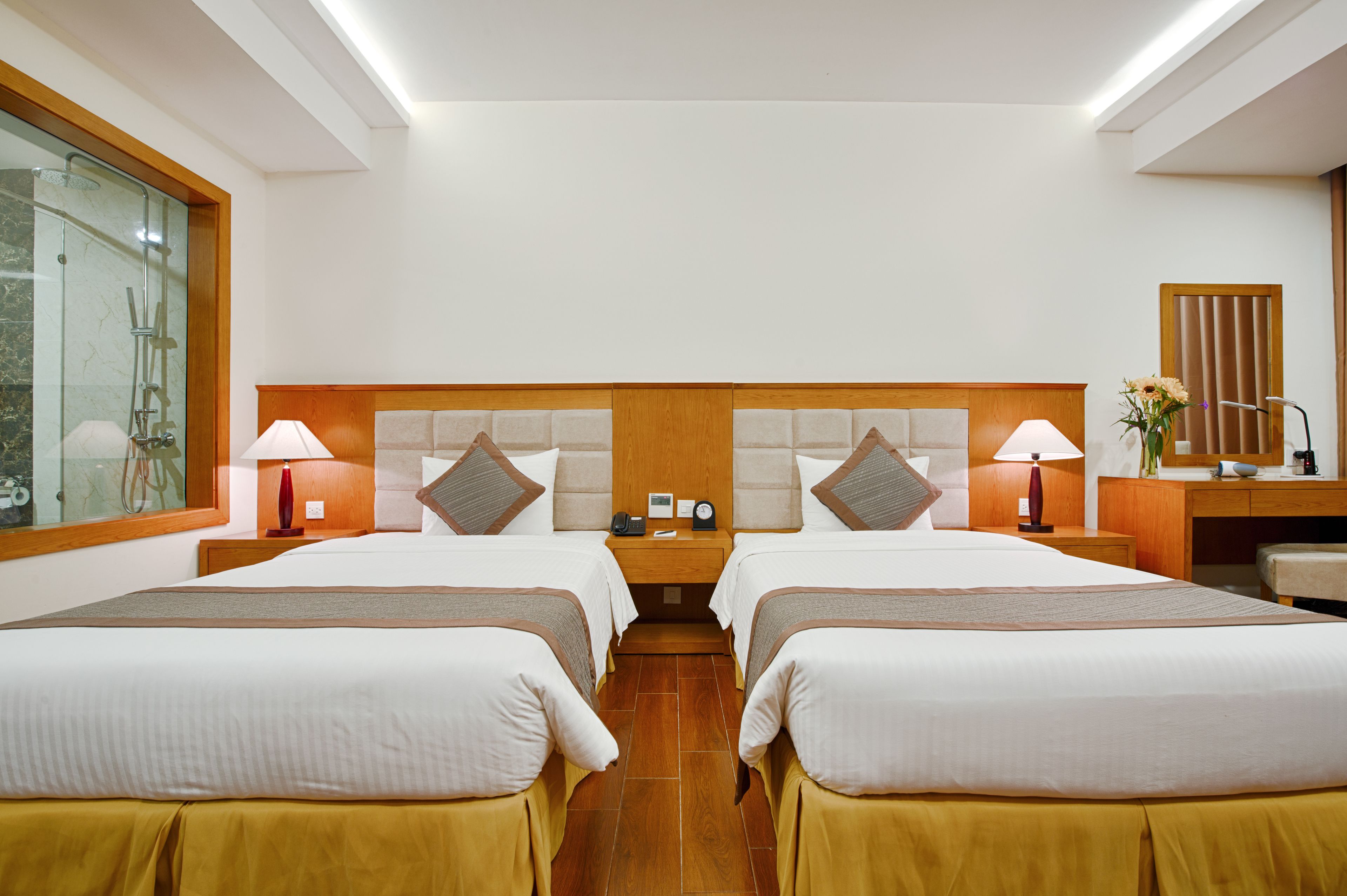 Quarto de hotel com duas camas, cabeceiras de madeira, luminárias de cabeceira, mesa de apoio e uma janela grande.