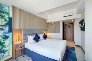 Quarto de hotel moderno com uma cama grande, uma mesinha de cabeceira, ar condicionado fixado na parede e um grande quadro abstrato à esquerda.