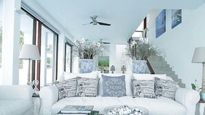 Uma sala de estar moderna com um sofá branco, almofadas decorativas, uma mesa de centro e uma escada ao fundo.