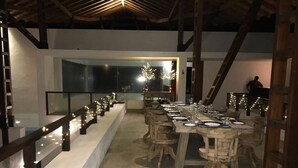 Uma longa mesa de jantar com cadeiras, posta para uma refeição, num espaço amplo com teto alto e vigas de madeira.