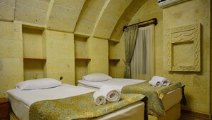 Quarto com duas camas, cada uma com colcha branca e manta estampada. Há uma mesa de cabeceira de madeira e uma pequena estante entre as camas. O quarto possui uma parede de pedra e uma janela com cortina.
