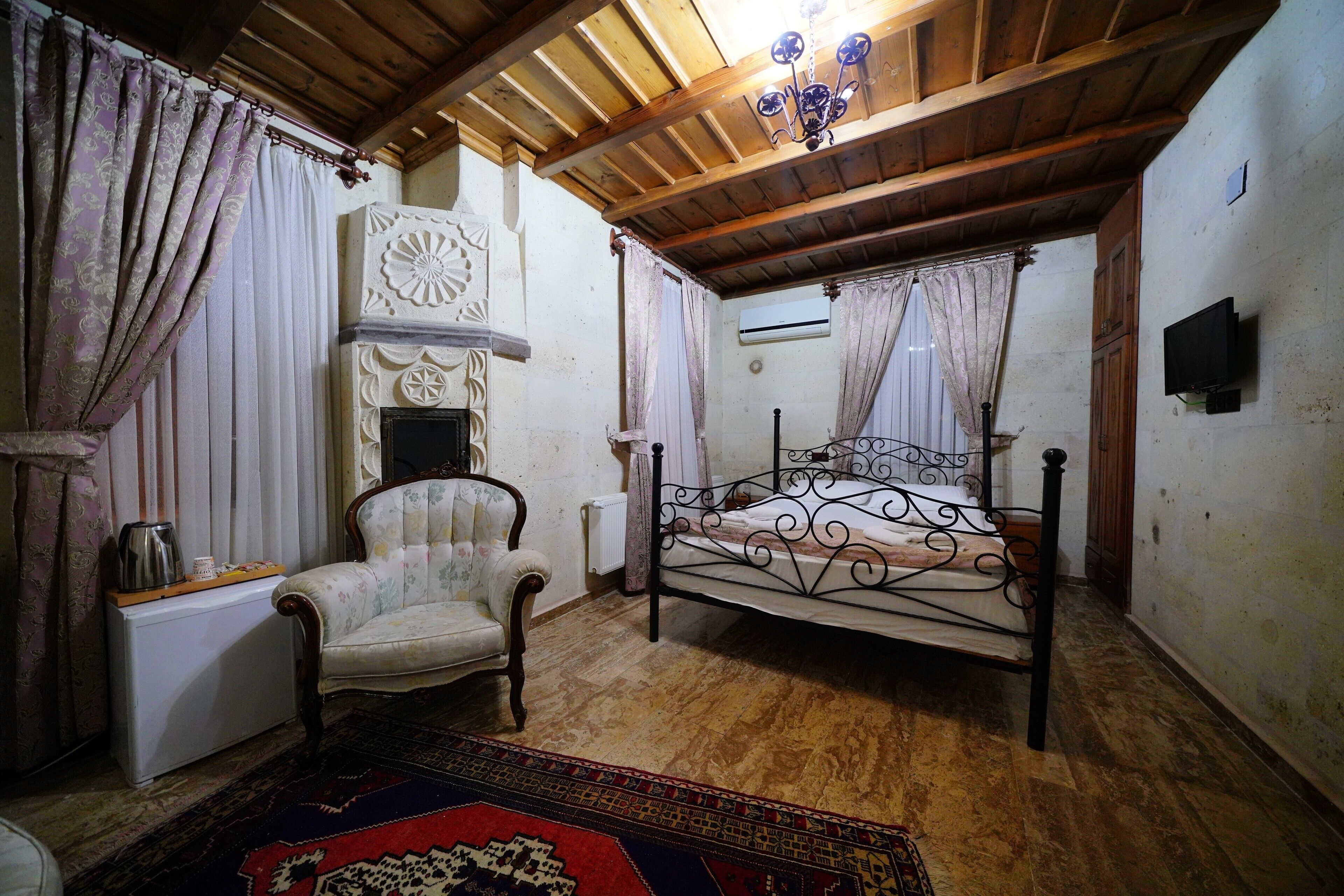 Quarto com teto de madeira, cama com estrutura de metal, cadeira, radiador e televisão na parede.
