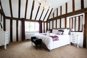 Quarto com vigas de madeira aparentes, uma cama grande com roupa de cama branca e detalhes roxos, uma mesa de cabeceira, um aparador e uma janela com cortinas.