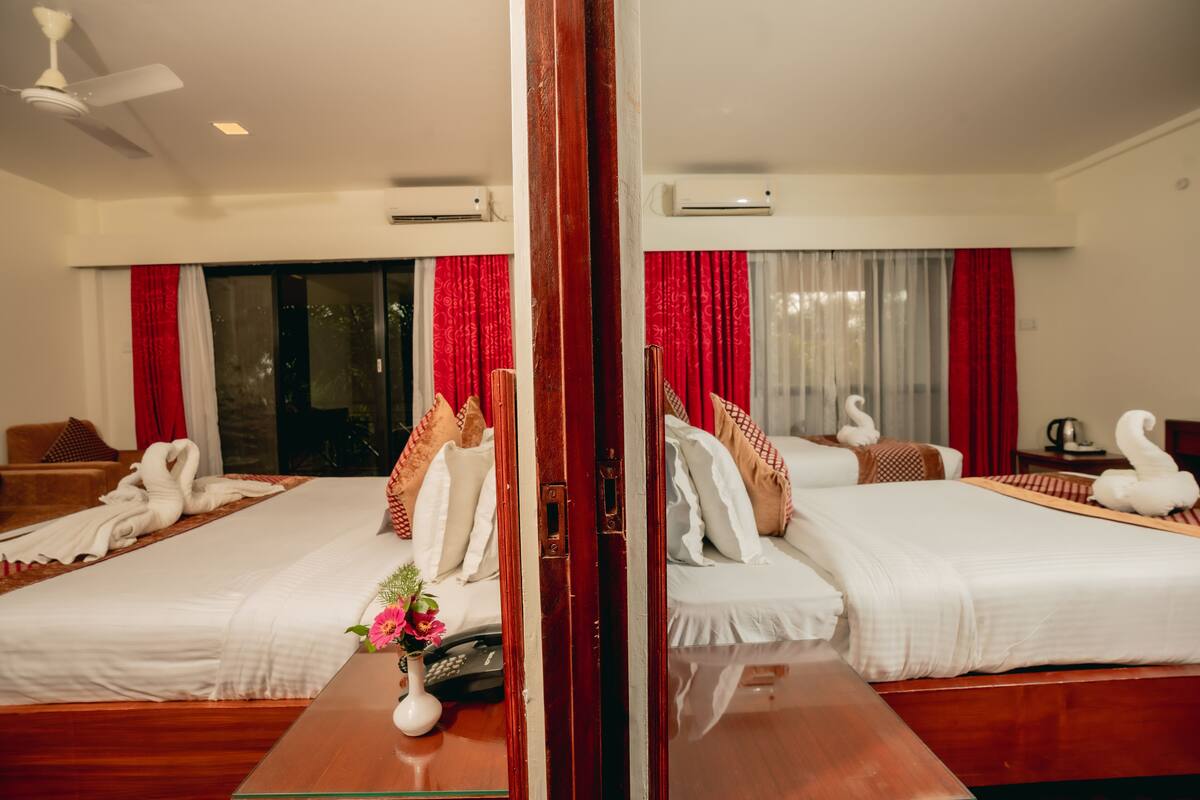 Quarto de hotel com duas camas, piso de madeira, espelho, um vaso com flores e cortinas vermelhas.