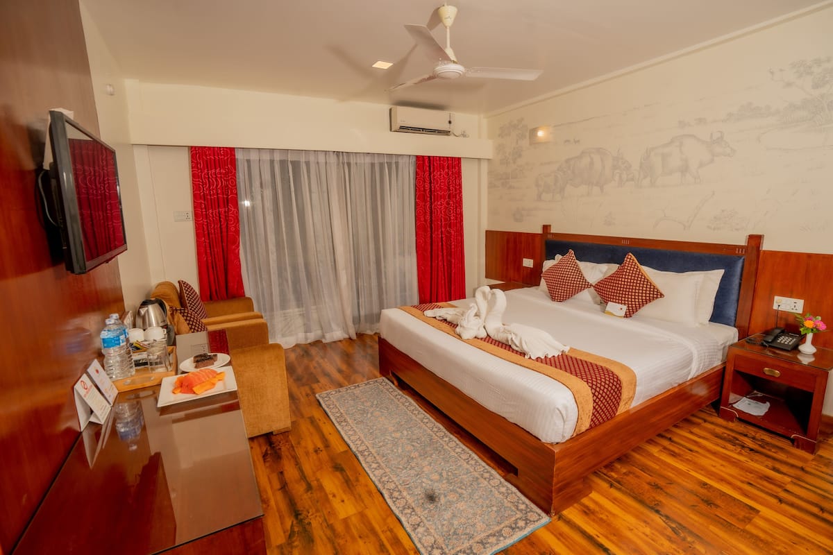 Quarto de hotel com cama de madeira, uma escrivaninha com telefone, uma mesinha com garrafa de água e cortina vermelha e branca.